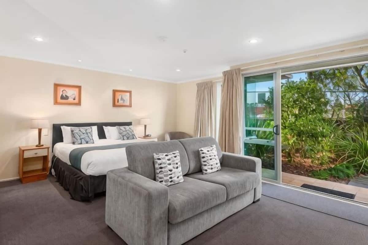 Quarto com cama, sofá cinzento, mesa de cabeceira e vista para o verde através de uma porta de vidro deslizante.