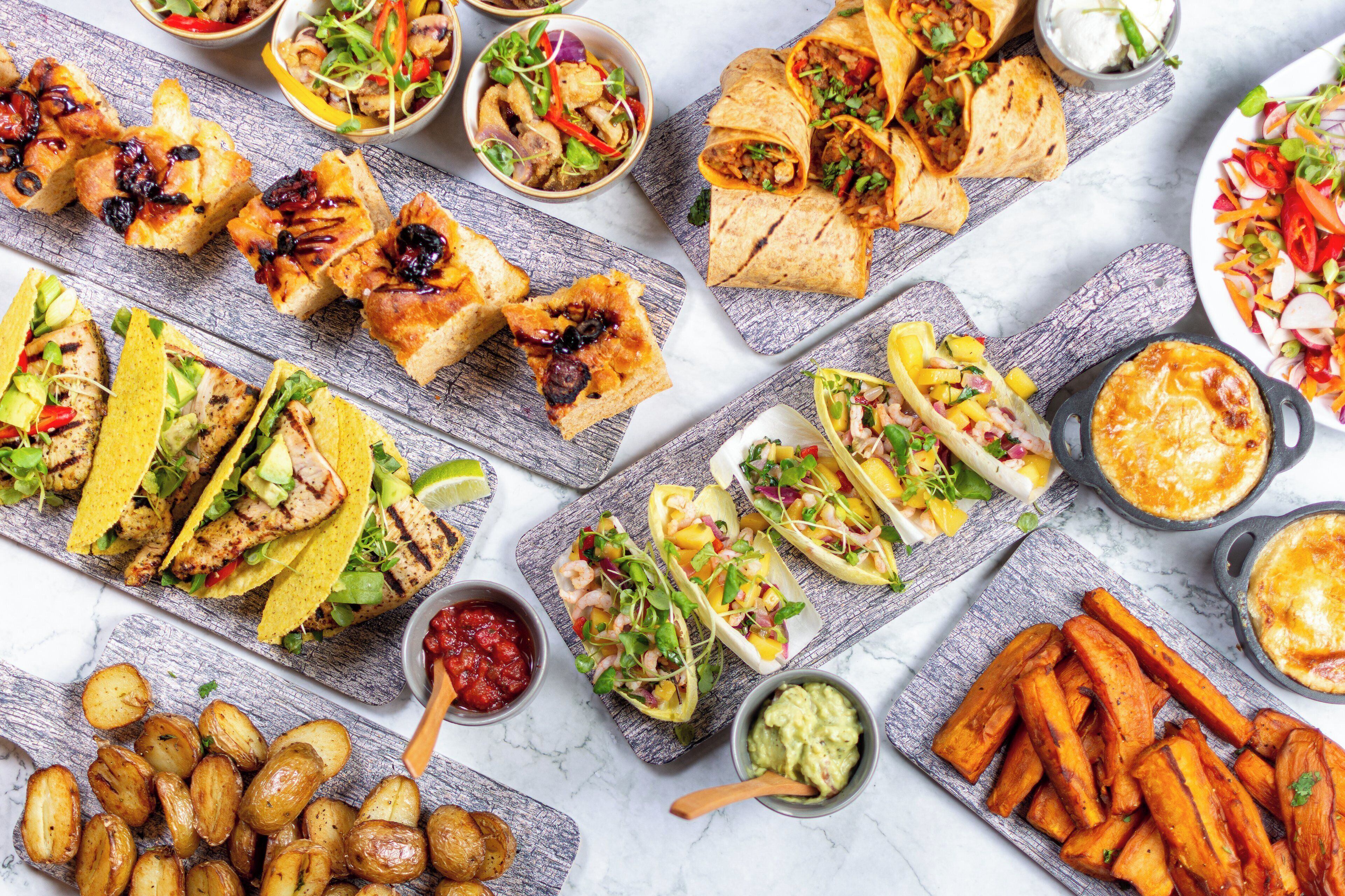Diversos pratos, incluindo tacos, burritos, frango grelhado e batatas assadas, indicando uma variedade de opções para a refeição.