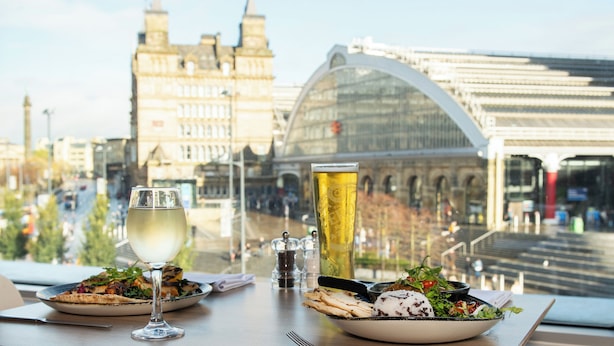 Mesa com refeição e bebidas, com vista para paisagem urbana com edifícios históricos e uma ponte moderna.