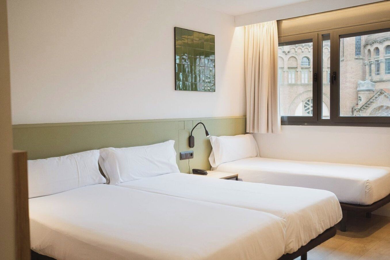 Quarto de hotel com duas camas, uma mesa de cabeceira, uma luminária e janela com vista para um edifício.