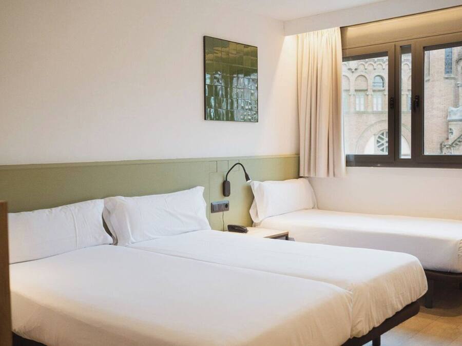 Quarto de hotel com duas camas, uma mesa de cabeceira, uma luminária e janela com vista para um edifício.