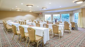 Sala de conferências com várias mesas e cadeiras, piso coberto de carpete e amplas janelas com cortinas.