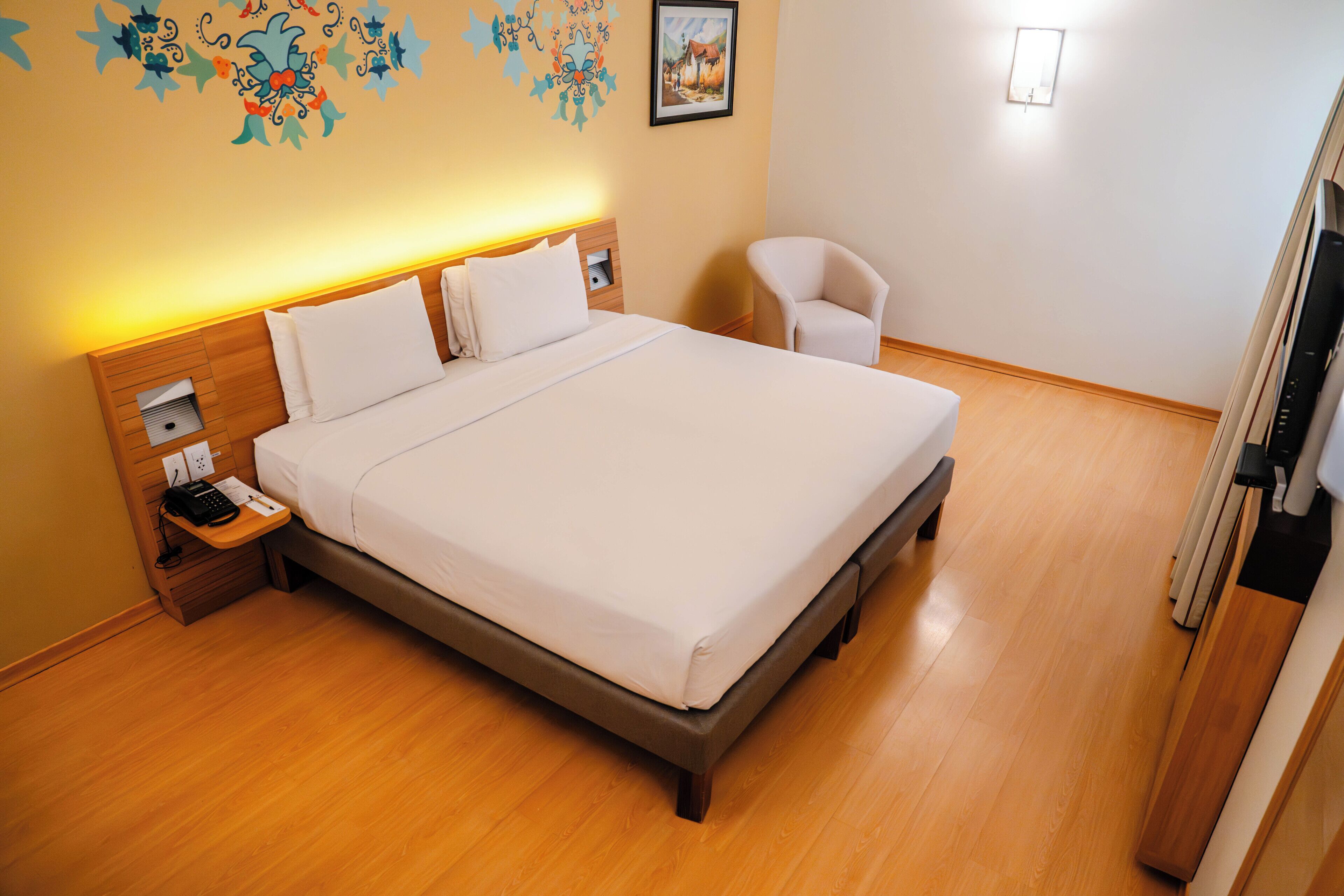 Cama bem arrumada com cabeceira de madeira, mesa de cabeceira com abajur e uma cadeira em quarto de hotel com piso de madeira e decoração na parede.