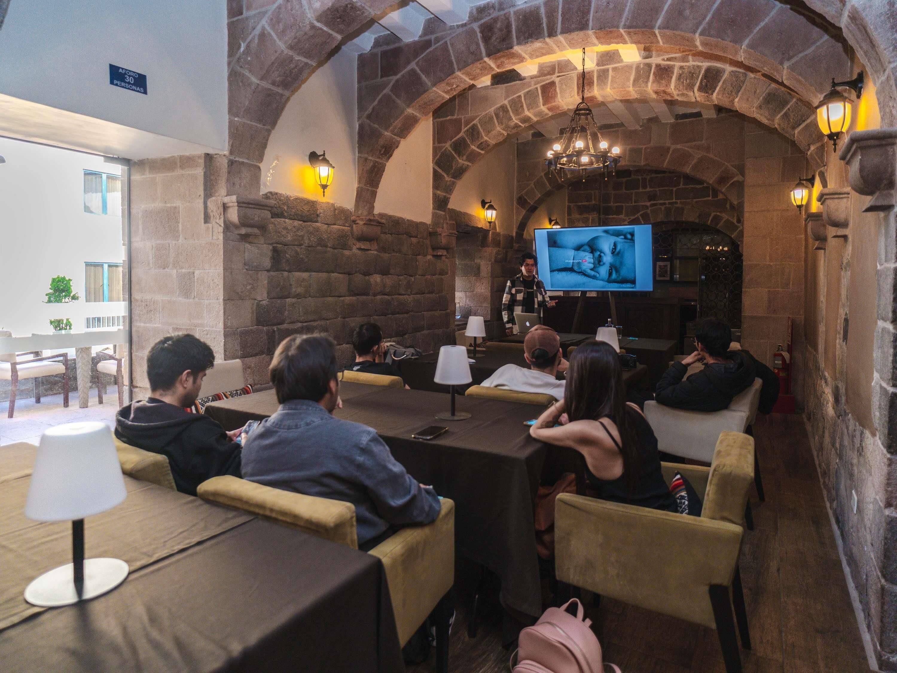Um grupo de pessoas sentadas em uma sala com paredes de pedra, mesas, cadeiras, uma tela de televisão e luminárias fixadas na parede.