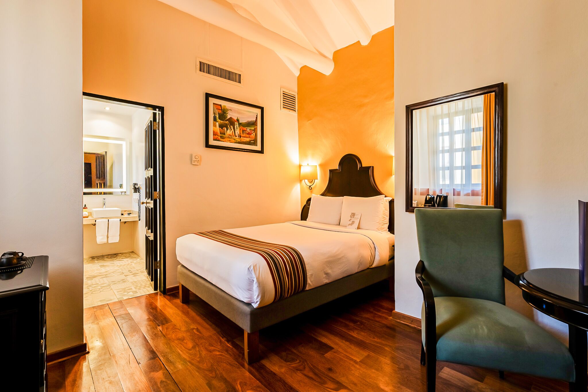 Quarto de hotel com cama, poltrona, escrivaninha e banheiro visível por uma porta aberta.