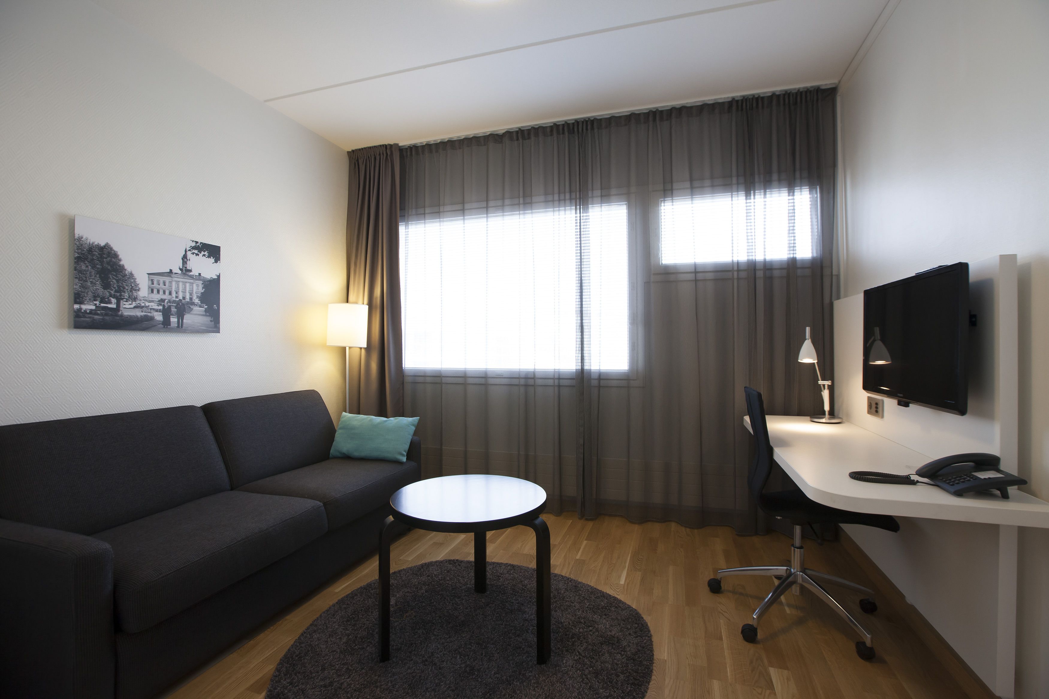 Quarto de hotel moderno com sofá escuro, mesa redonda pequena, escrivaninha com cadeira, televisão e janela com cortinas translúcidas.