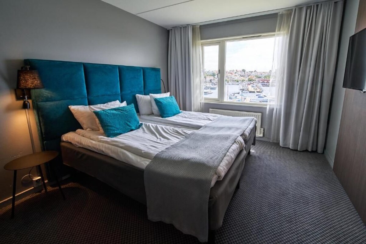 Quarto de hotel com uma cama grande, mesa de cabeceira, abajur e vista para a cidade pela janela.