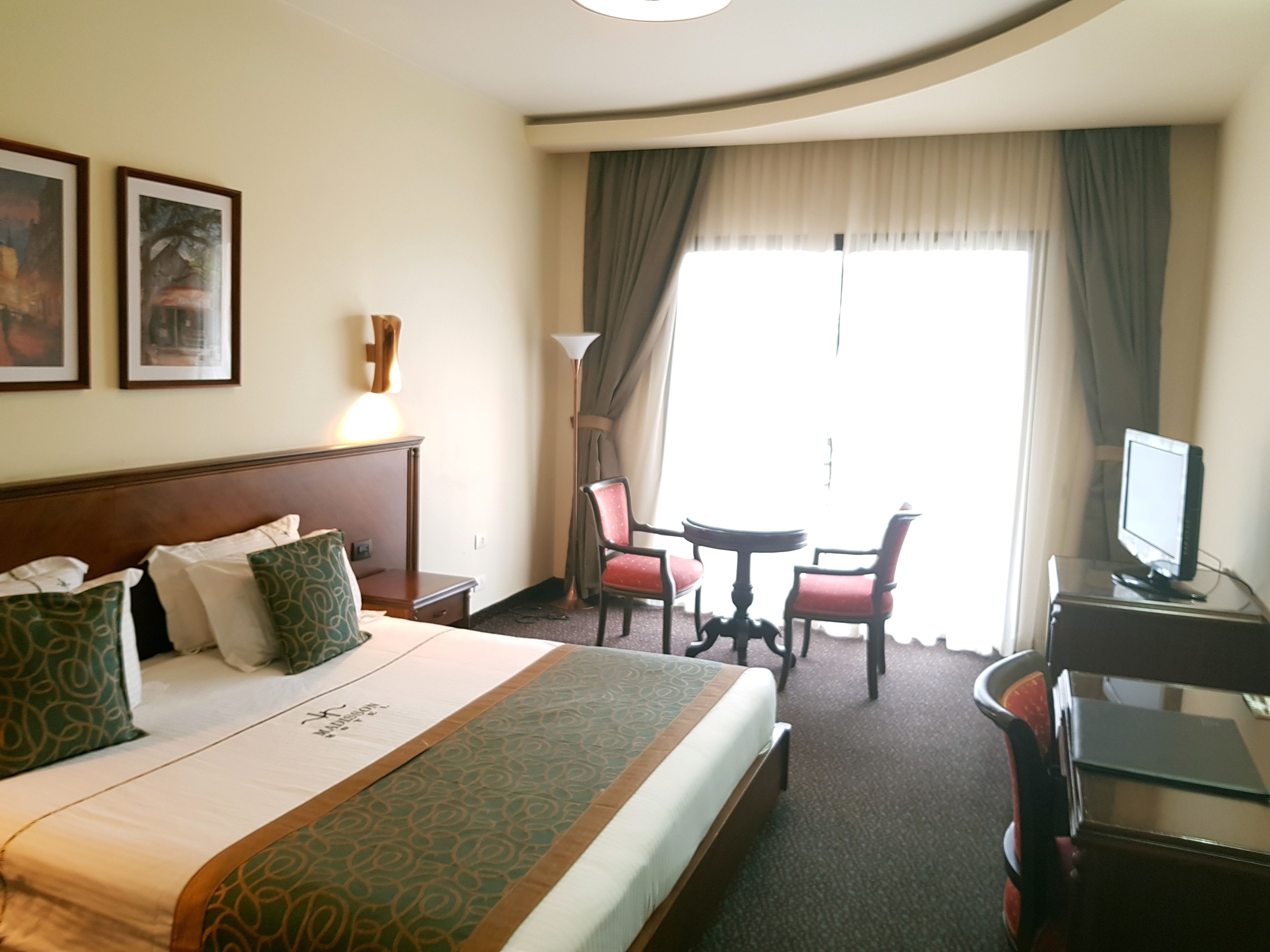 Quarto de hotel com cama, escrivaninha com computador, duas cadeiras, abajur e janela com cortinas.