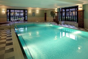 Piscina interna com água cristalina, piso ladrilhado e algumas cadeiras de descanso.