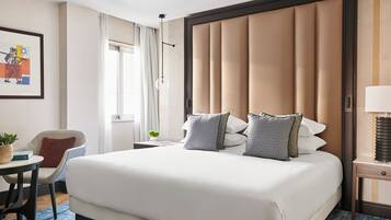 Quarto de hotel com uma cama grande, uma cadeira, uma mesinha com uma planta, uma luminária e uma janela com cortinas.