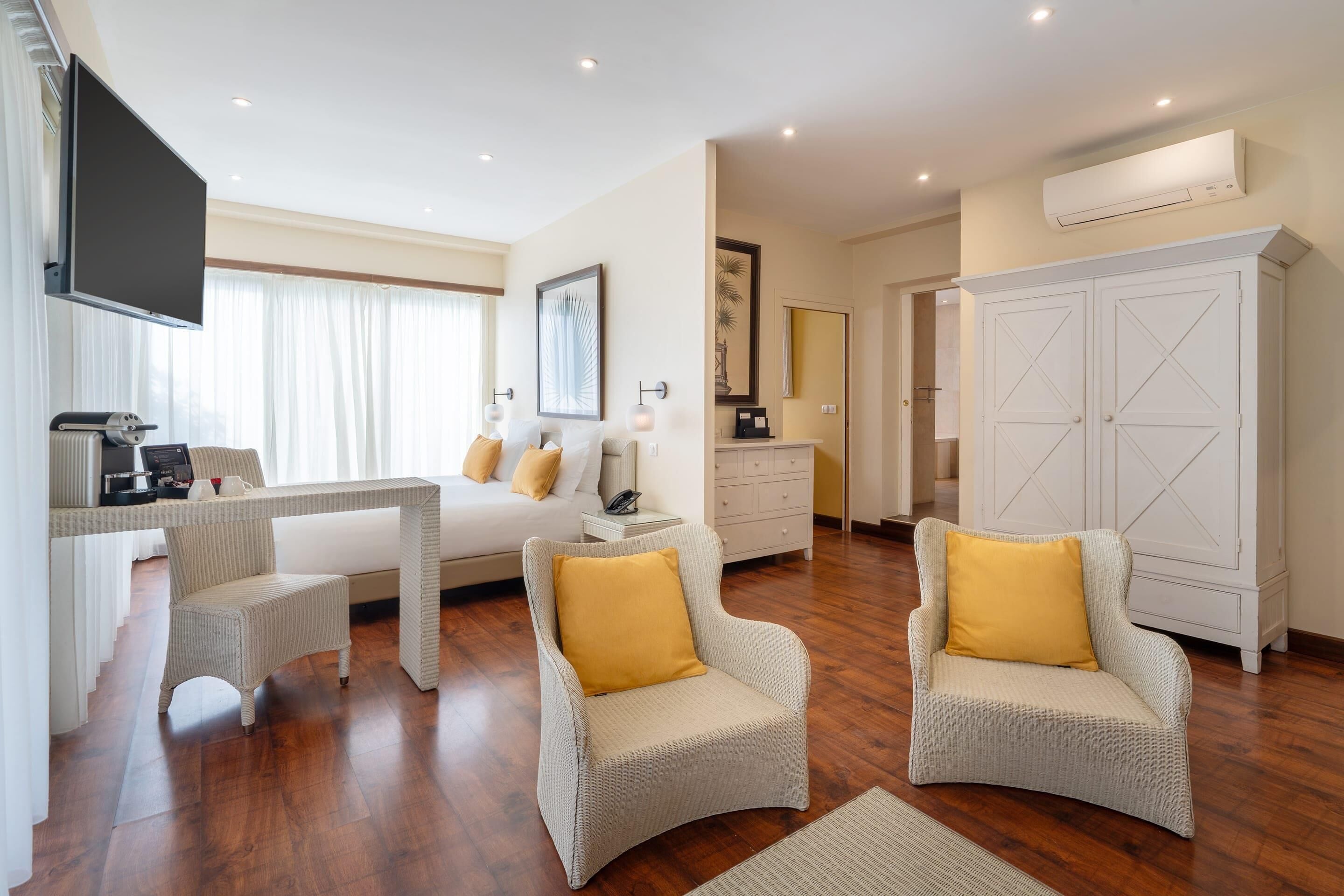 Quarto de hotel com cama, duas poltronas, escrivaninha, televisão e guarda-roupa.