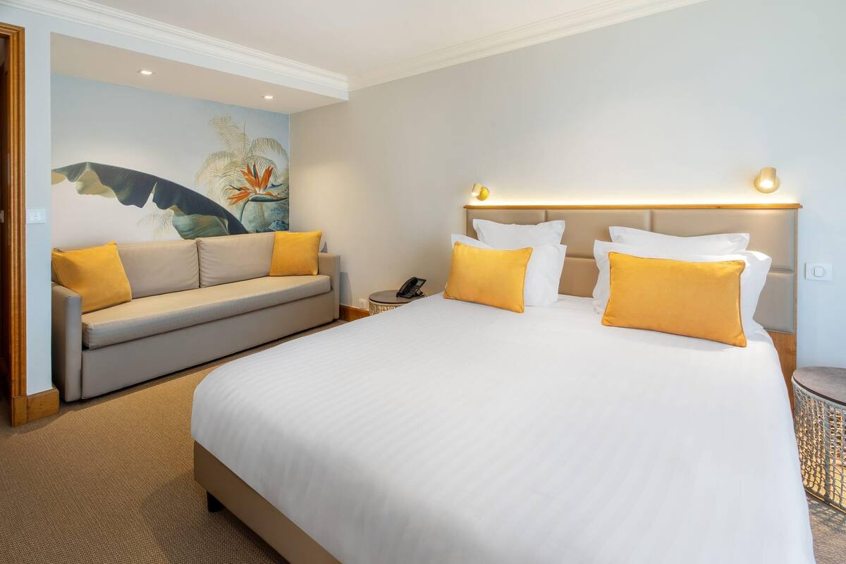 Quarto de hotel com uma cama grande, um sofá, um mural na parede e uma mesa de cabeceira.