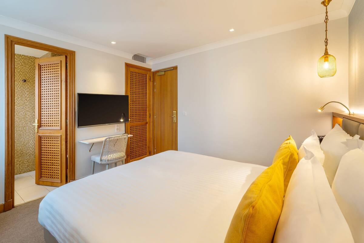 Quarto de hotel com uma cama, uma escrivaninha com cadeira, televisão e uma porta com detalhe em madeira treliçada.