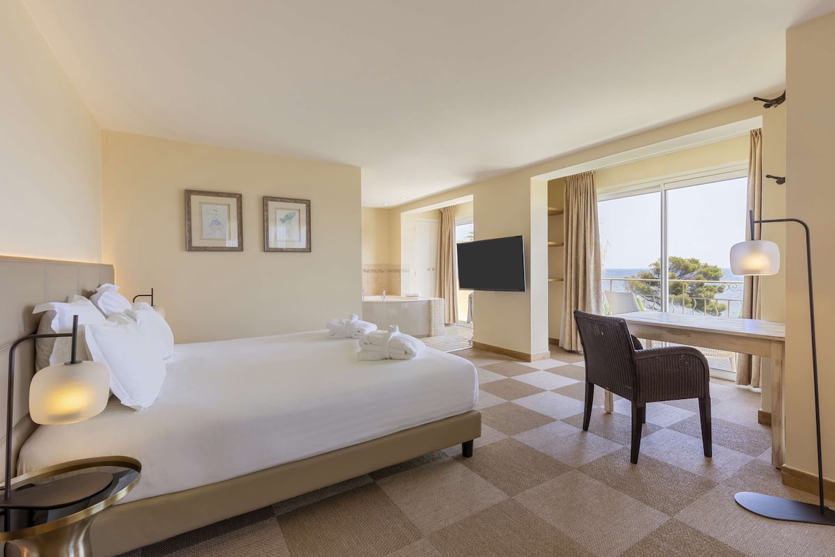 Quarto de hotel com uma cama grande, uma escrivaninha, uma cadeira, uma TV e vista para o mar.