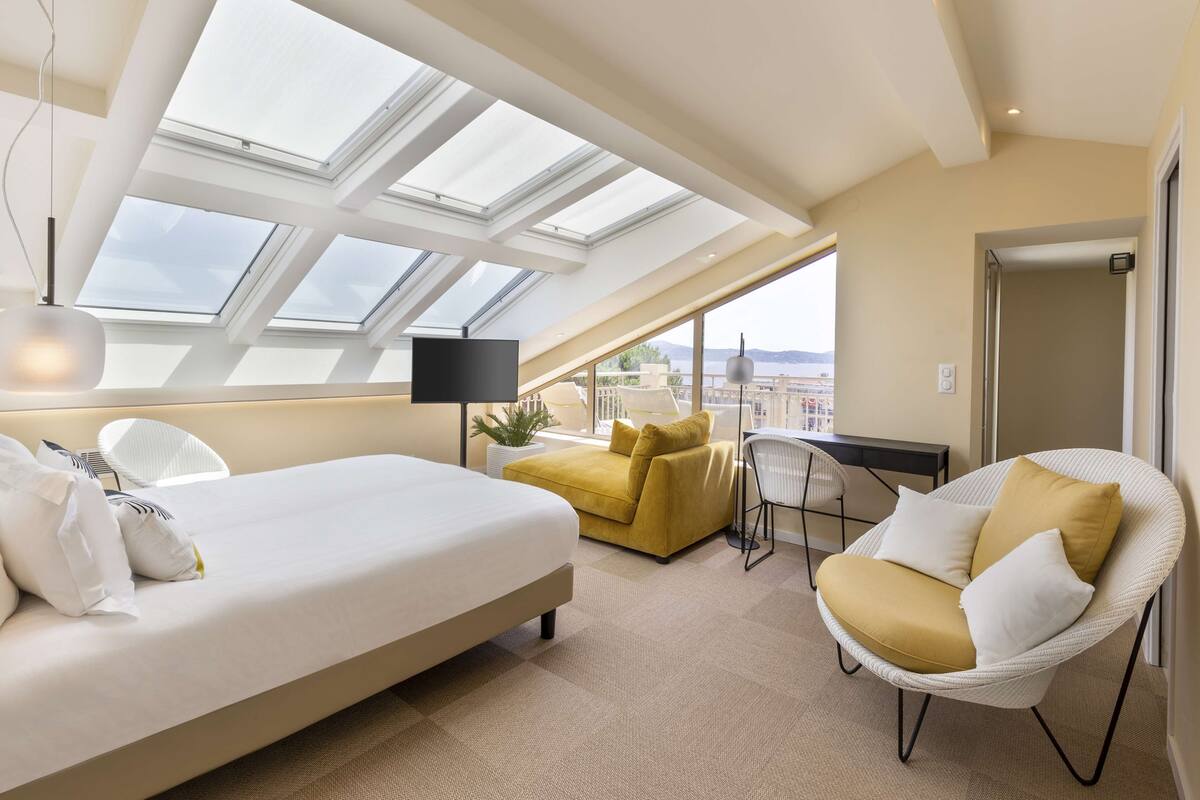 Quarto moderno com clarabóia, uma cama, um sofá amarelo e uma cadeira branca.