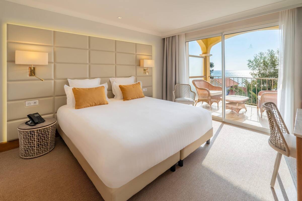 Quarto de hotel com uma cama grande, abajures, telefone, cadeira de vime e varanda com mobiliário exterior.