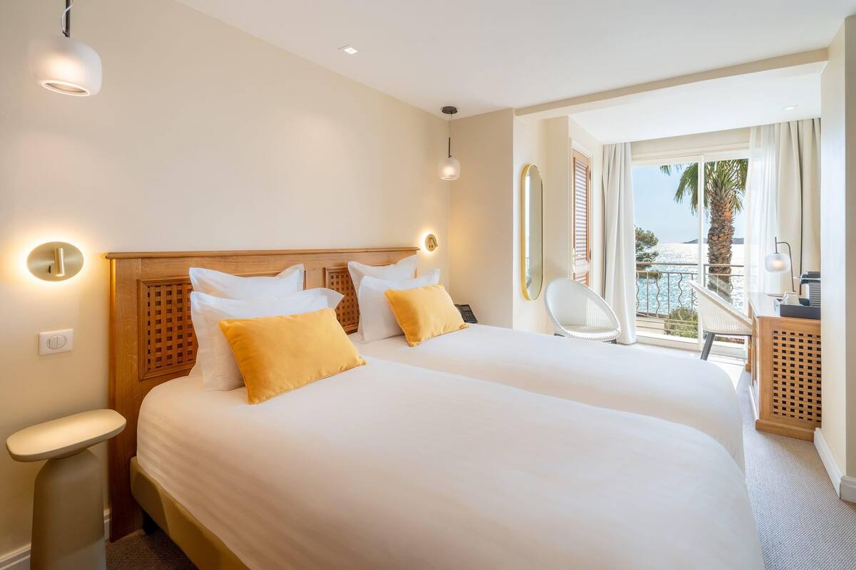 Quarto de hotel com duas camas, varanda e vista, e secretária com cadeira.