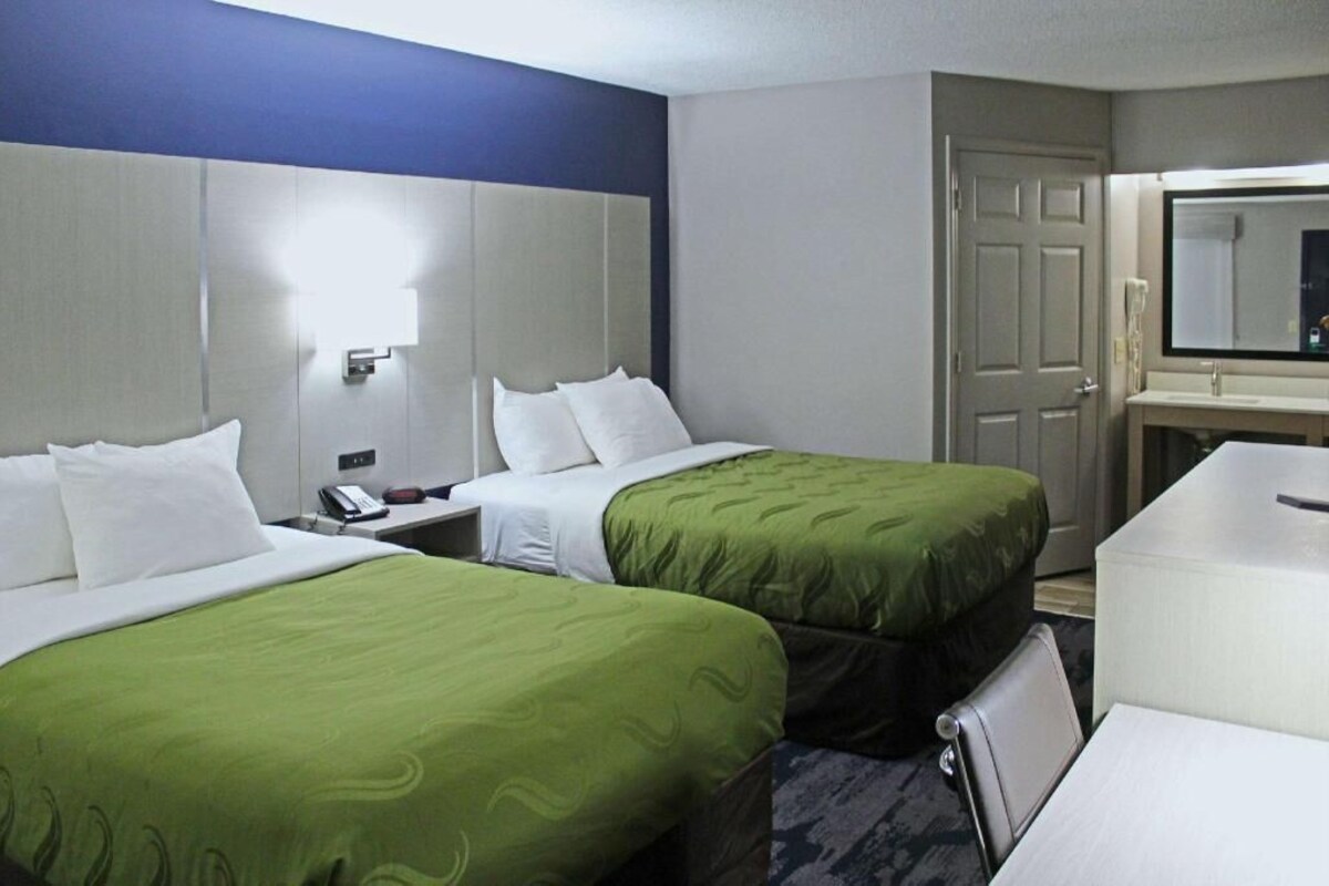 Quarto de hotel com duas camas, uma escrivaninha, uma cadeira, um espelho e uma porta.