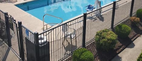 Piscina retangular rodeada por uma cerca de metal preto, com espreguiçadeiras e uma pequena mesa de um lado.