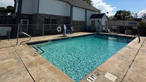Piscina com deck ladrilhado, edifício com fachada branca e detalhes em pedra, e céu azul límpido.