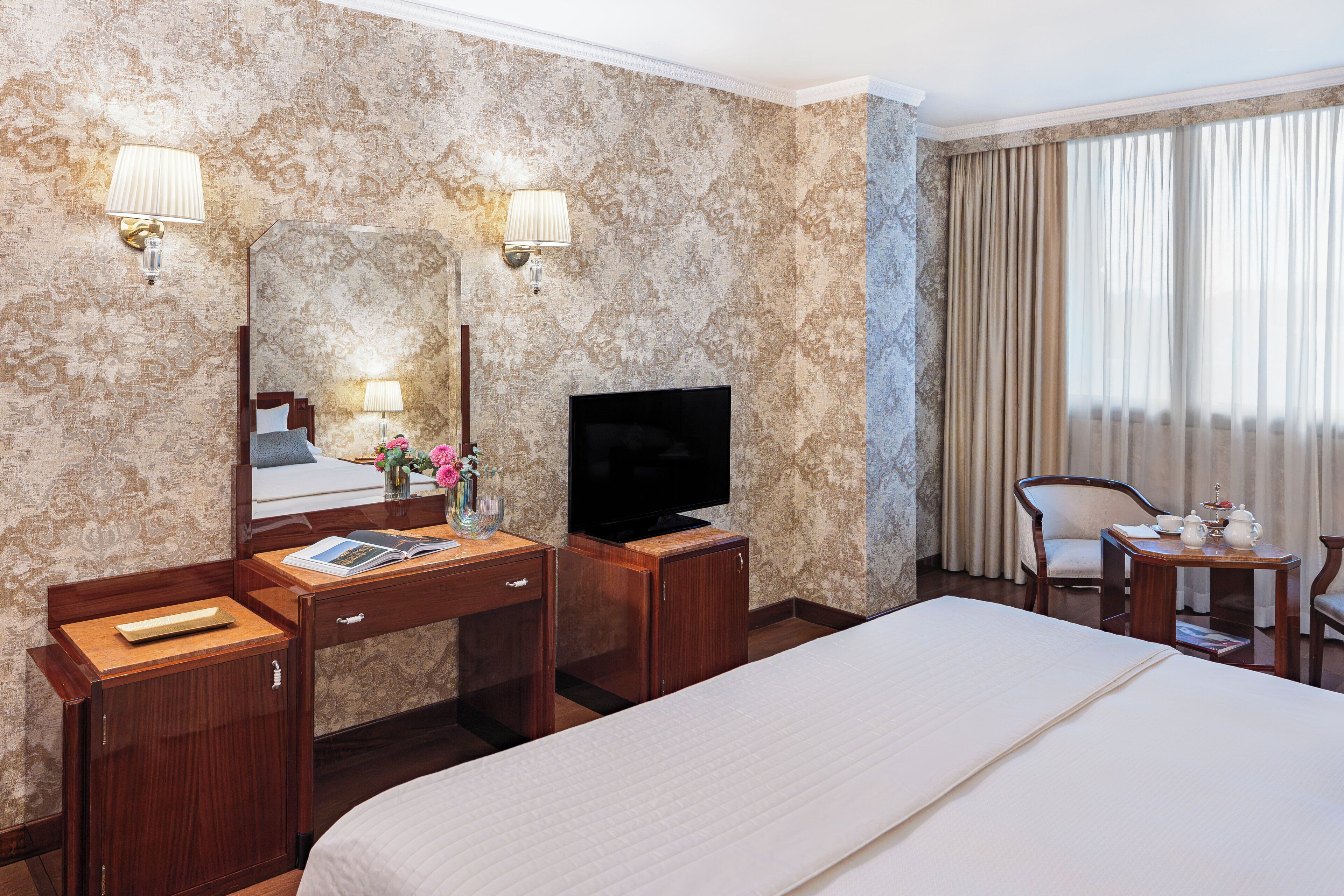 Quarto de hotel com uma cama grande, uma escrivaninha com televisão, um espelho e uma janela com cortinas.