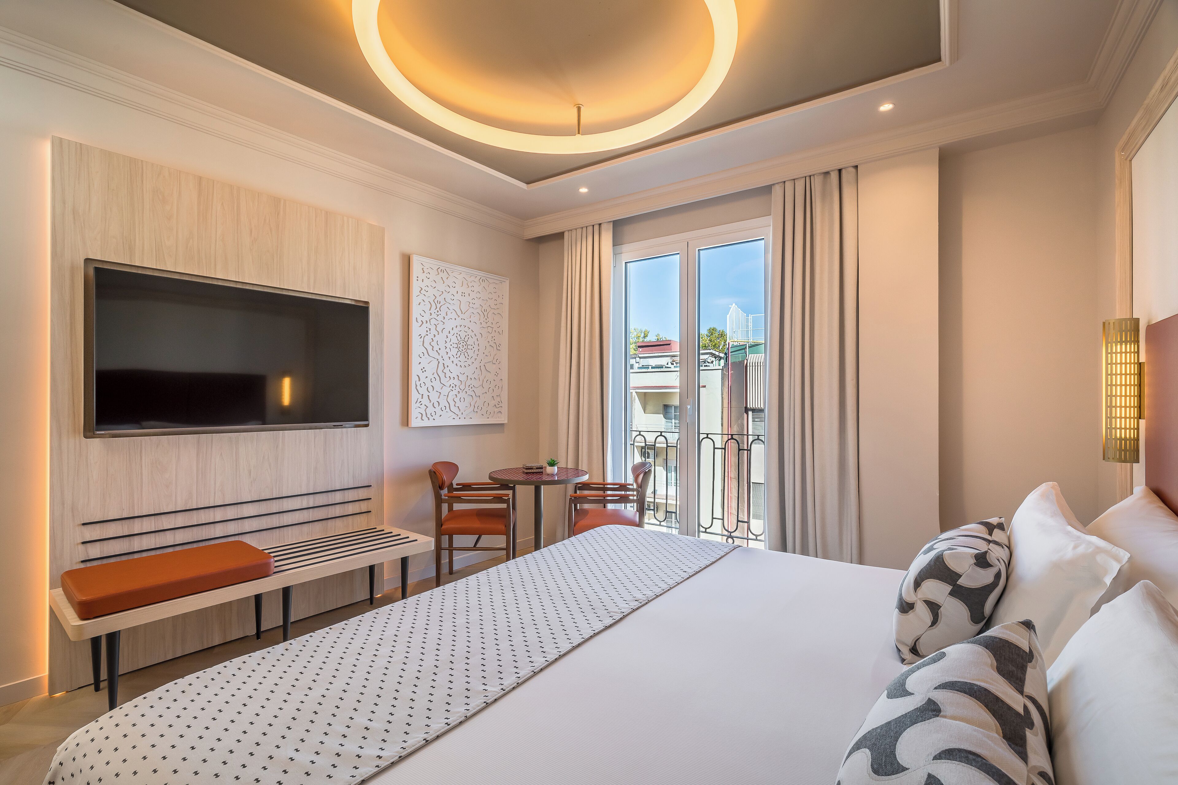 Quarto de hotel com uma cama grande, televisão na parede, uma mesa pequena com cadeiras e uma varanda com cortinas.