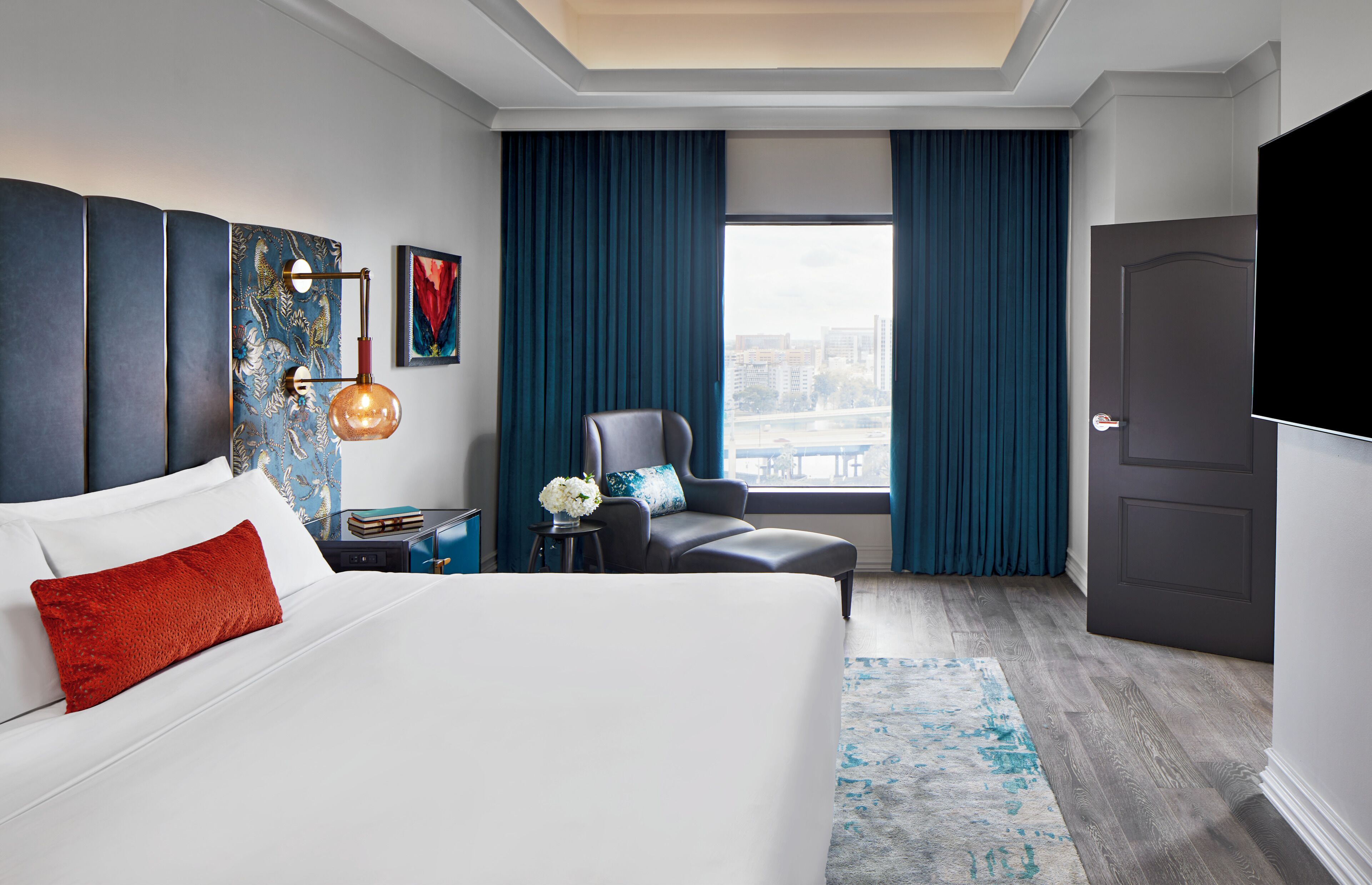 Quarto de hotel com cama, escrivaninha, cadeira, televisão e uma janela grande com cortinas.