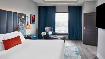 Quarto de hotel com cama, escrivaninha, cadeira, televisão e uma janela grande com cortinas.