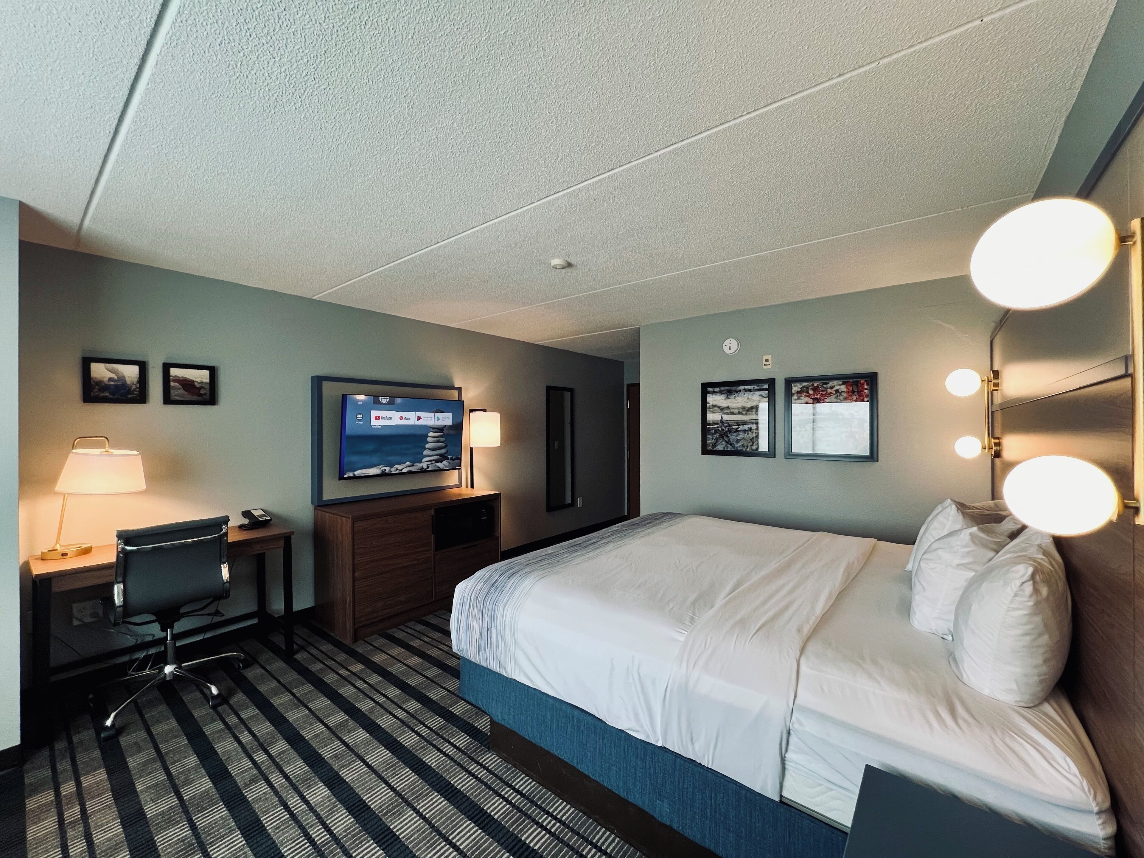 Quarto de hotel com cama, escrivaninha, cadeira, televisão e luminárias fixadas na parede.