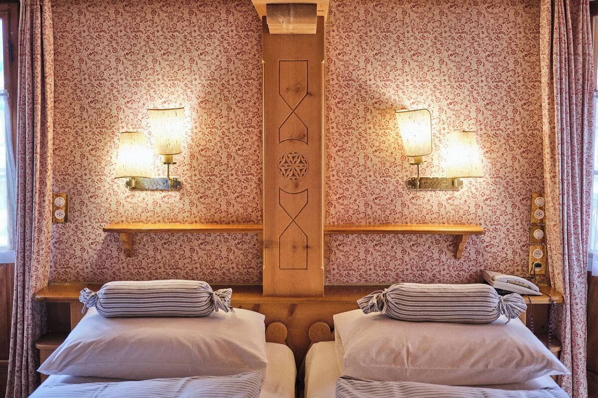 Quarto com duas camas, cada uma com travesseiro e mesa de cabeceira. Há luminárias fixadas na parede em ambos os lados do quarto.