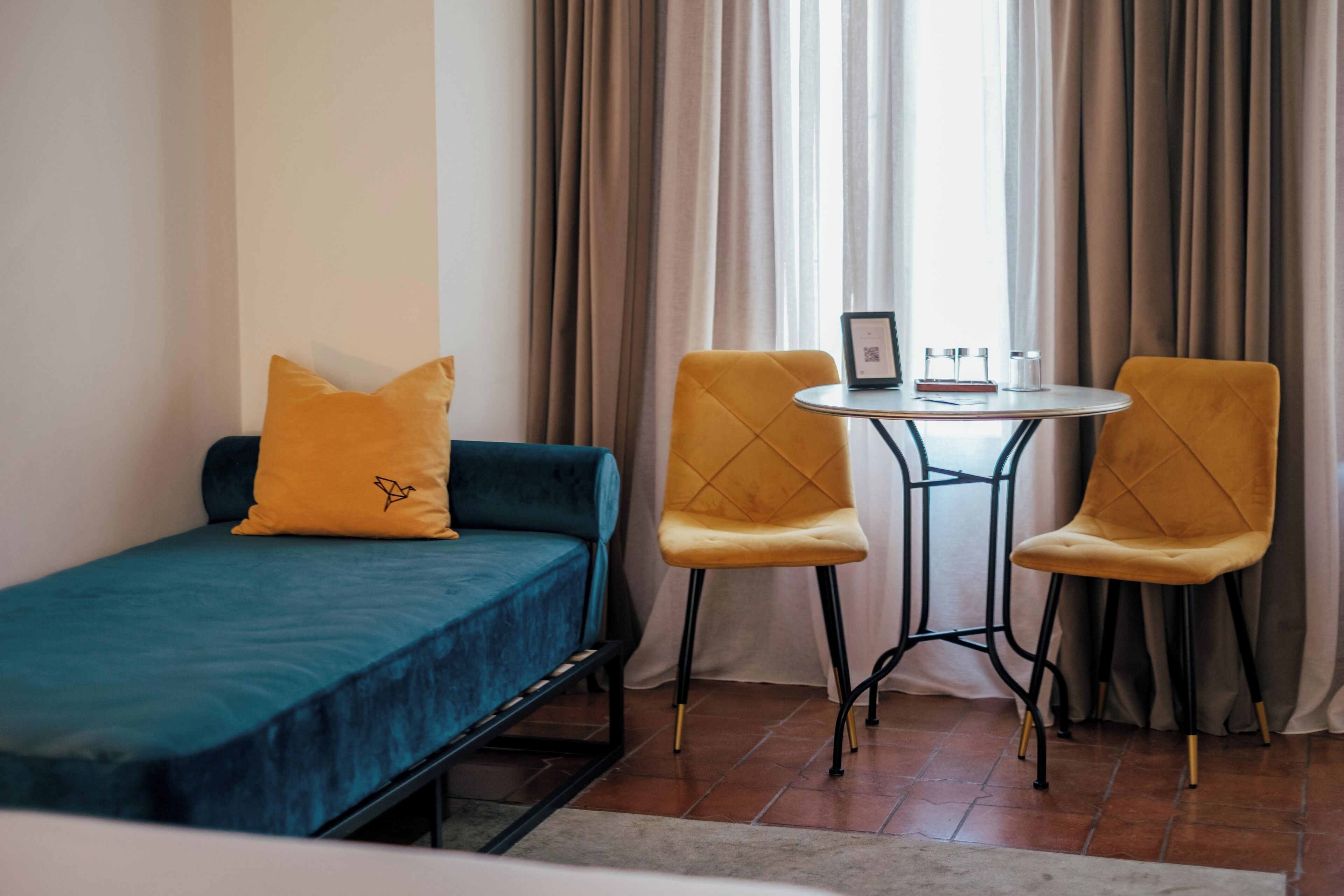 Quarto com sofá azul-turquesa, uma mesinha com cadeiras e uma janela com cortinas.