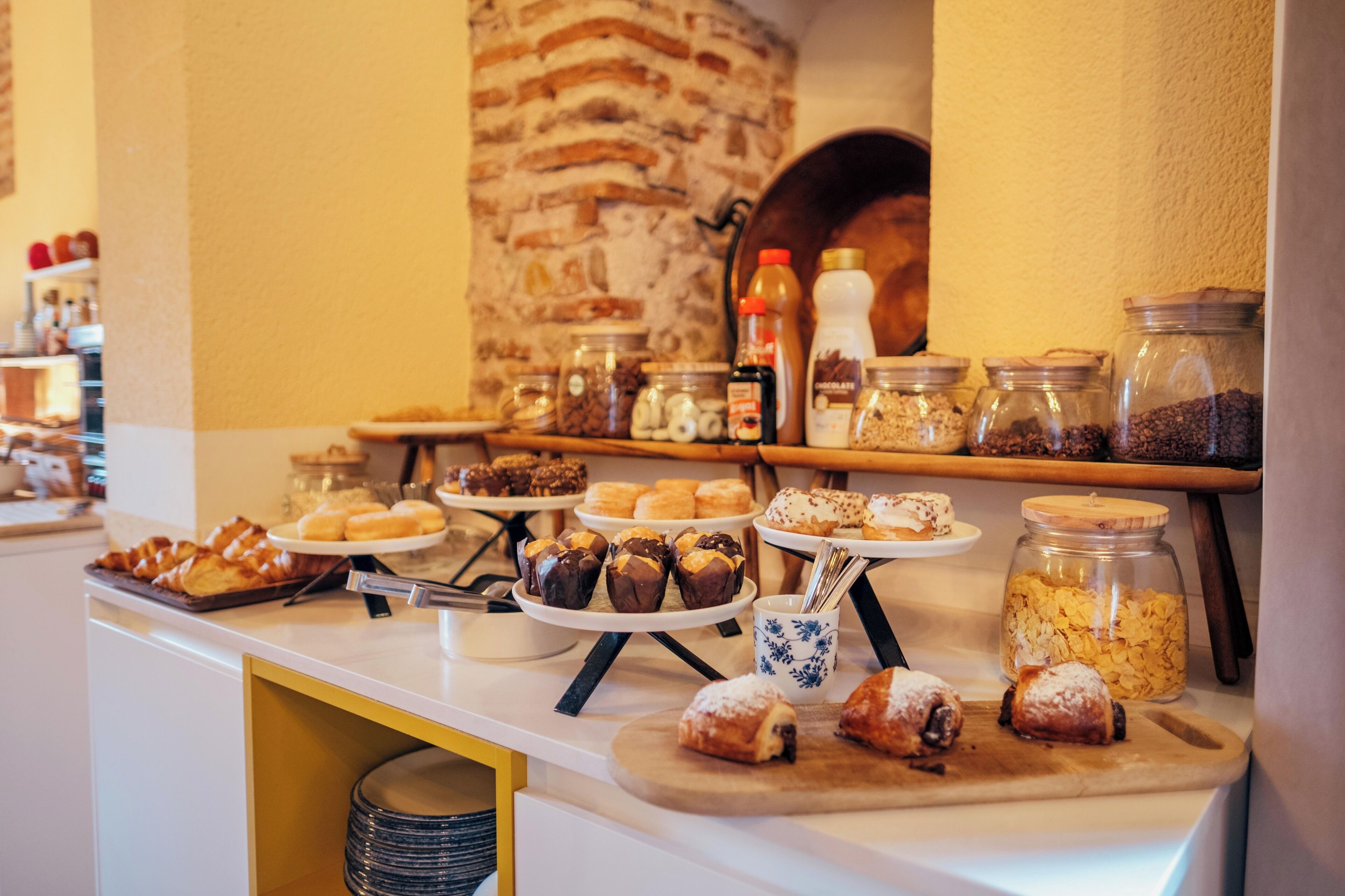 Buffet com diversos pastéis e sobremesas, incluindo croissants e muffins, exposto em um balcão com parede de tijolos ao fundo.