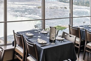 Mesa de restaurante posta com pratos brancos, talheres e copos, um suporte para menu e vista para um rio através de amplas janelas.