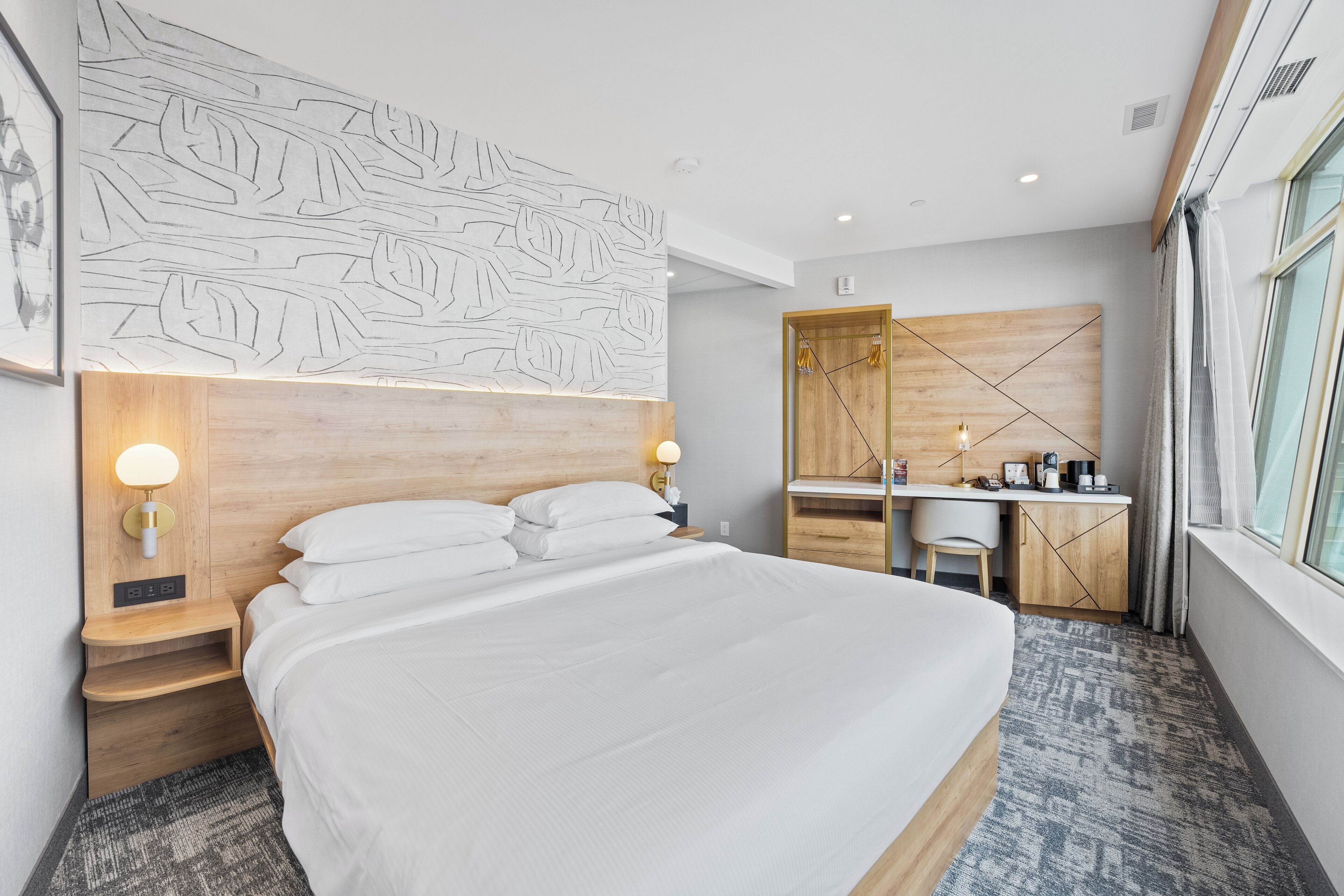 Quarto de hotel moderno com uma cama grande, mesas de cabeceira com abajures, um painel de cama de madeira, uma escrivaninha com cadeira e uma janela ampla.