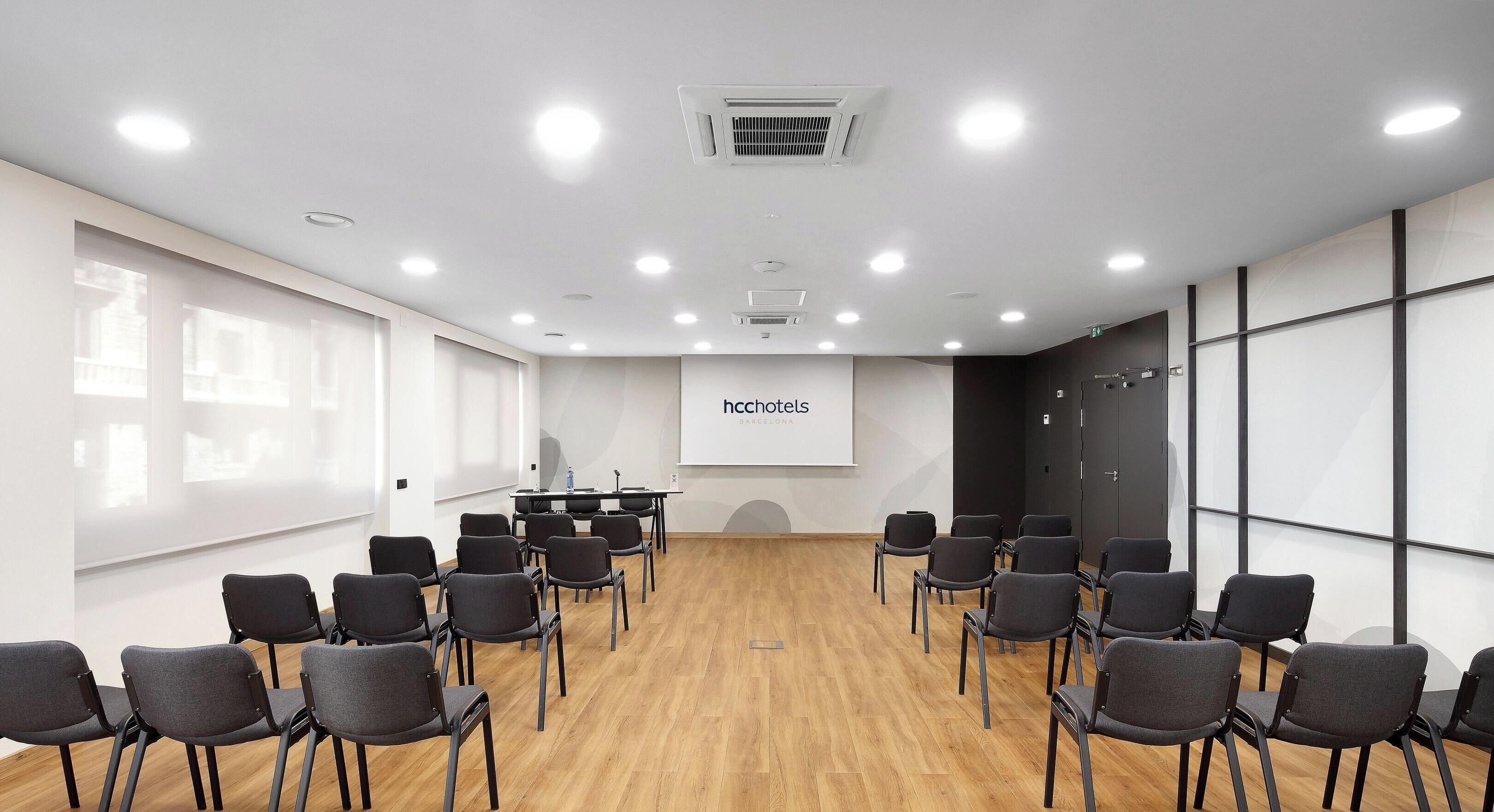 Sala de conferências com tela de projetor exibindo 'hchotels Barcelona', fileiras de cadeiras e dutos de ventilação no teto.