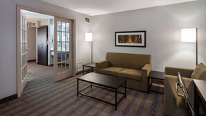 Quarto de hotel com sofá, mesa de centro, mesa lateral e luminária. Há uma porta que dá para um banheiro e um corredor.