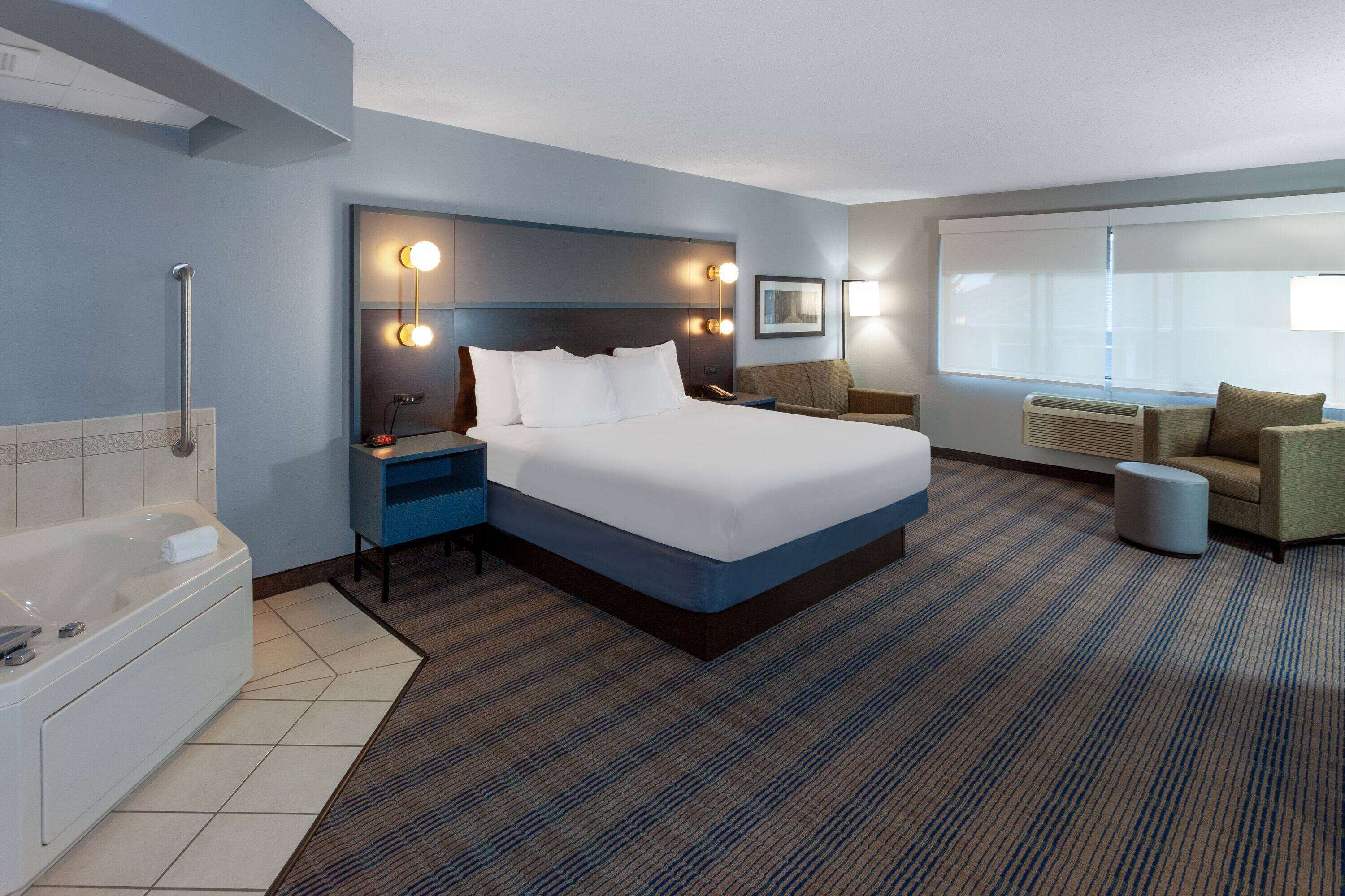 Quarto de hotel com cama grande, área de estar separada, banheira e janela com persianas.