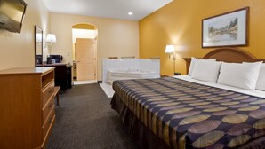 Quarto de hotel com cama, escrivaninha com computador, luminária de mesa, televisão, banheiro com banheira e um quadro na parede.