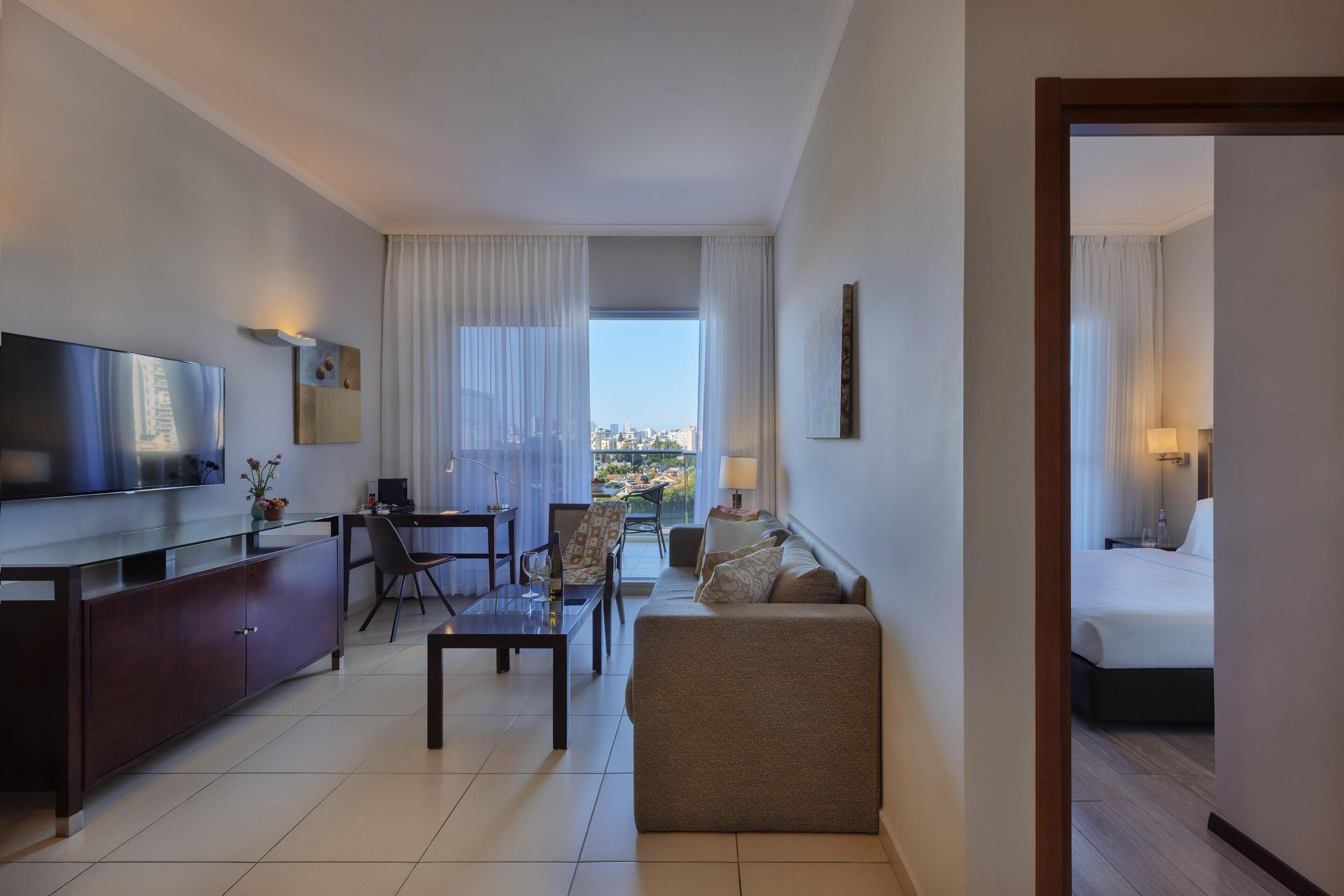 Quarto de hotel moderno com uma televisão de tela grande, um sofá, uma área de refeições e uma varanda com vista para a cidade.