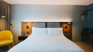 Quarto de hotel com uma cama grande, mesa de cabeceira com telefone, uma cadeira e uma escrivaninha.