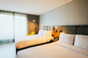 Quarto de hotel com duas camas, uma escrivaninha, uma cadeira e uma janela grande com cortinas.