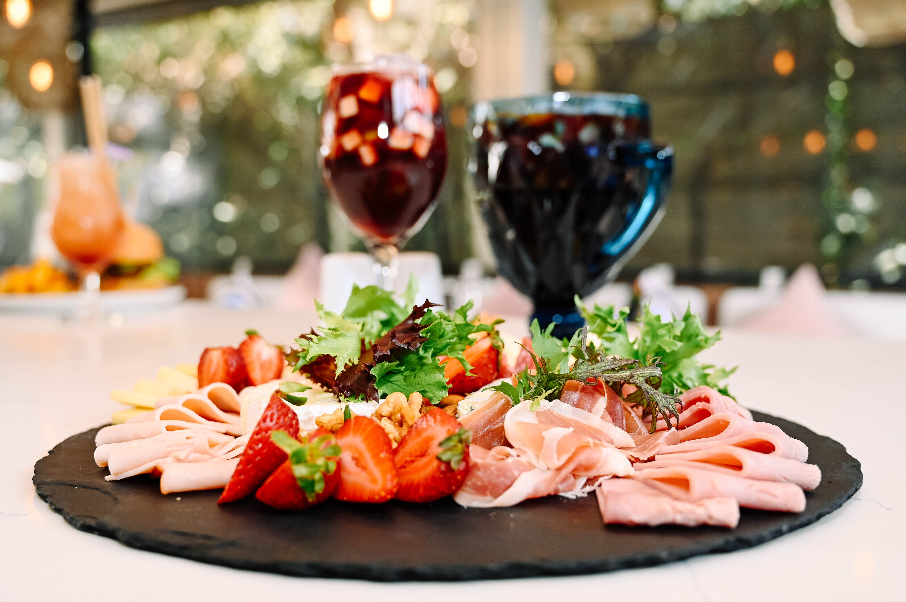 Tábua de frios com diversos embutidos, frutas e vegetais, acompanhada por dois copos de vinho tinto.