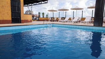 Piscina com espreguiçadeiras e guarda-sóis, cercada por um muro revestido e entrada de um edifício.