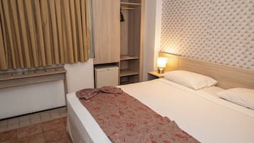 Quarto de hotel com cama, mesa de cabeceira com luminária, guarda-roupa e janela com cortinas.