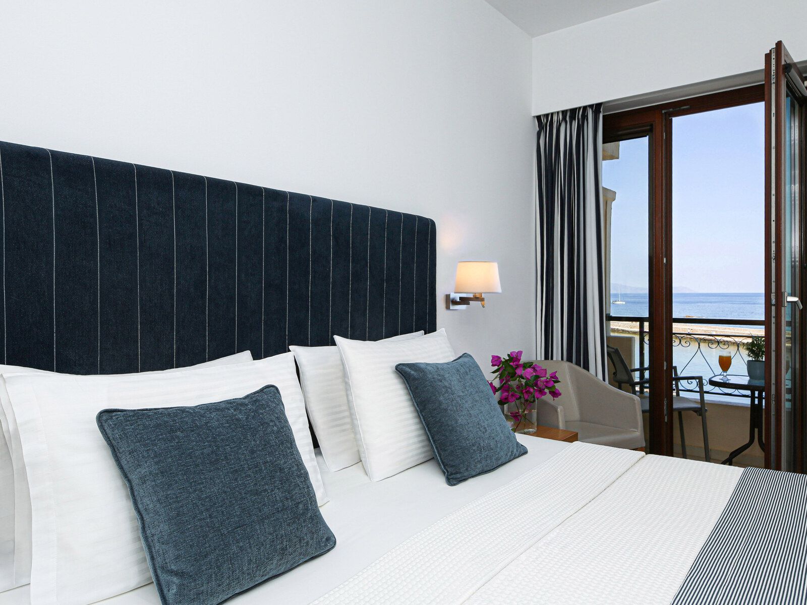 Quarto de hotel com uma cama grande, mesa de cabeceira com abajur, vista para o mar através de uma porta de varanda e uma planta em vaso.