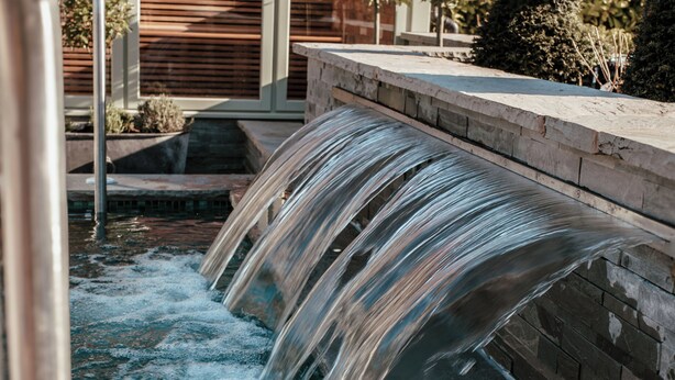 Piscina moderna ao ar livre com uma cascata, rodeada por um deck de madeira e uma área de estar coberta.