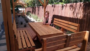 Um pátio externo de madeira com uma mesa e bancos, cercado por um muro de madeira e vegetação.