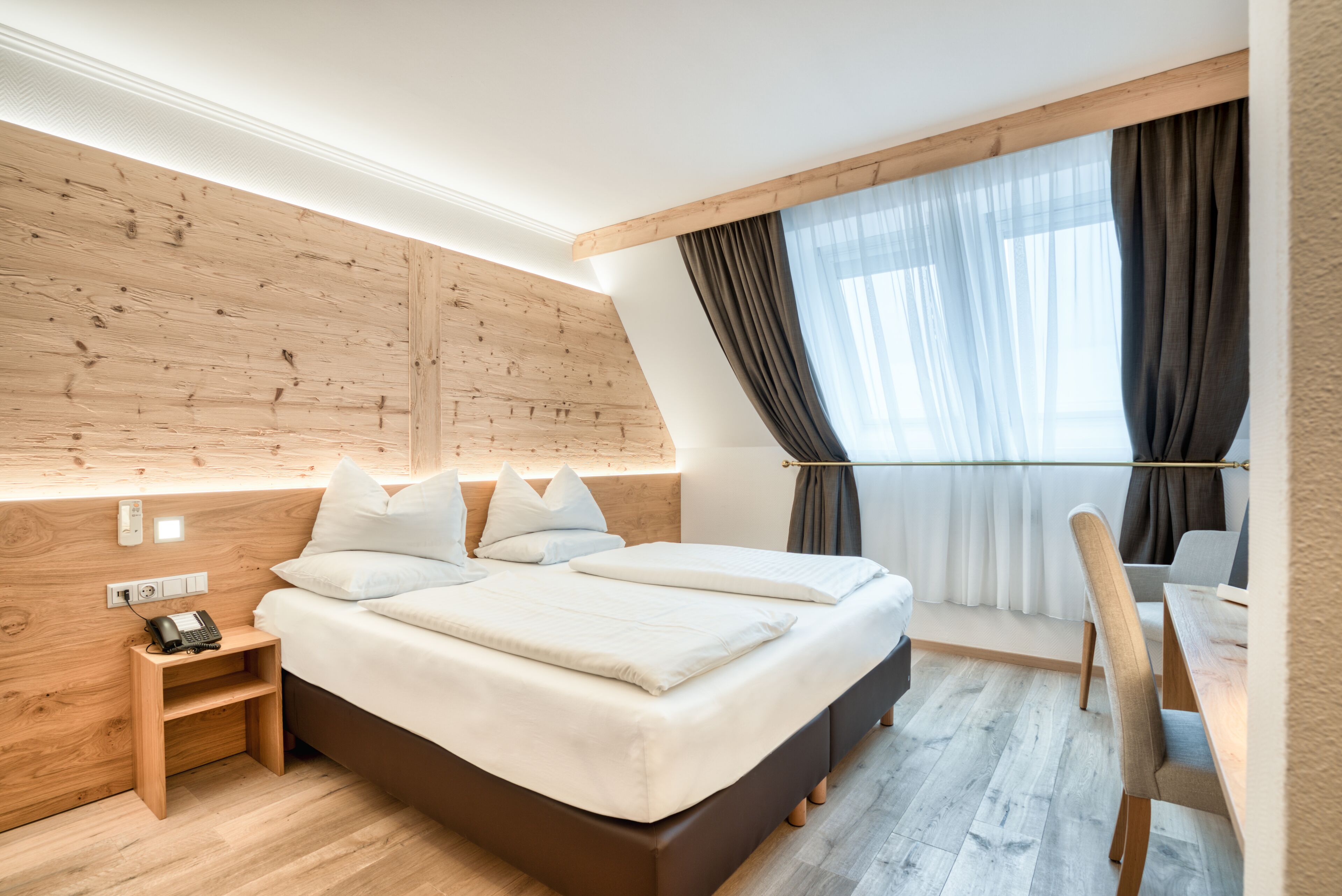 Quarto de hotel moderno com cabeceira de madeira, uma cama grande com roupa de cama branca, uma mesinha de cabeceira com telefone e vista da cidade através de cortinas leves.