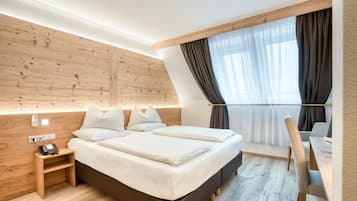 Quarto de hotel moderno com cabeceira de madeira, uma cama grande com roupa de cama branca, uma mesinha de cabeceira com telefone e vista da cidade através de cortinas leves.