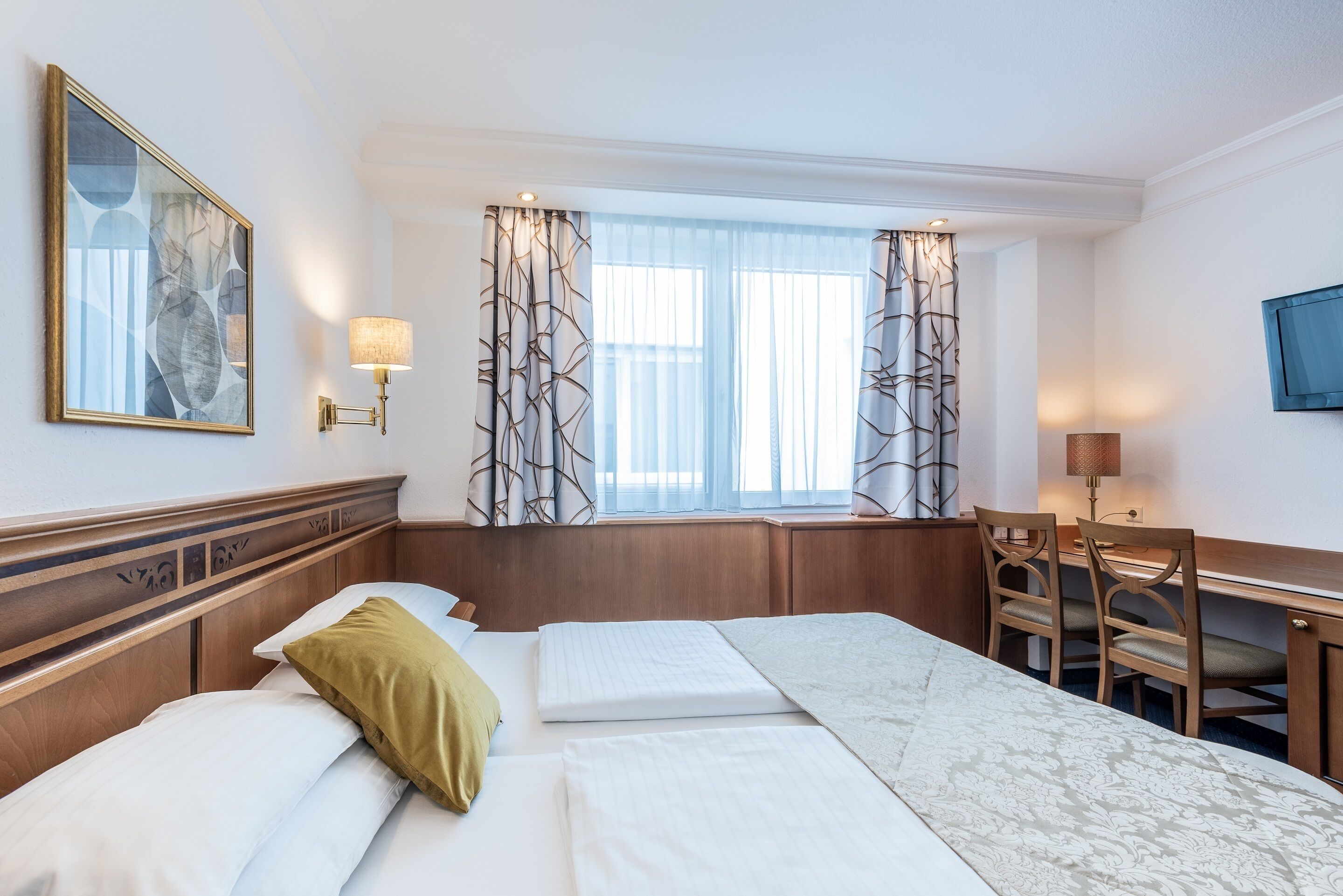 Quarto de hotel com uma cama grande, uma mesa com duas cadeiras, uma televisão na parede e uma janela com cortinas.